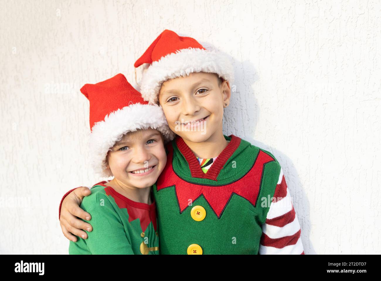 two smiling cute boys in Santa hats and elf costumes hug each other on ...