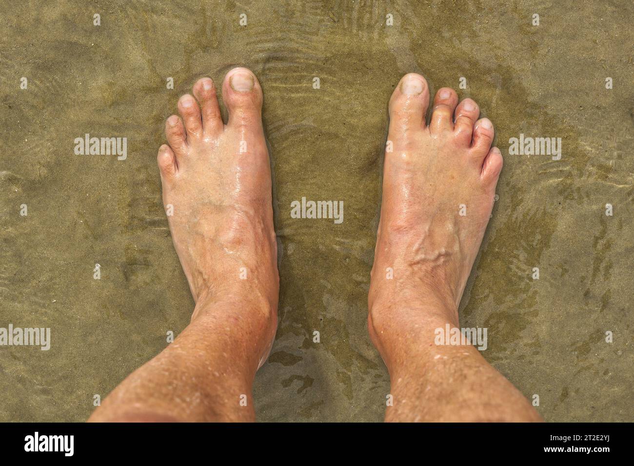 Pair of adult male feet on sandy beach with clear water washing over ...