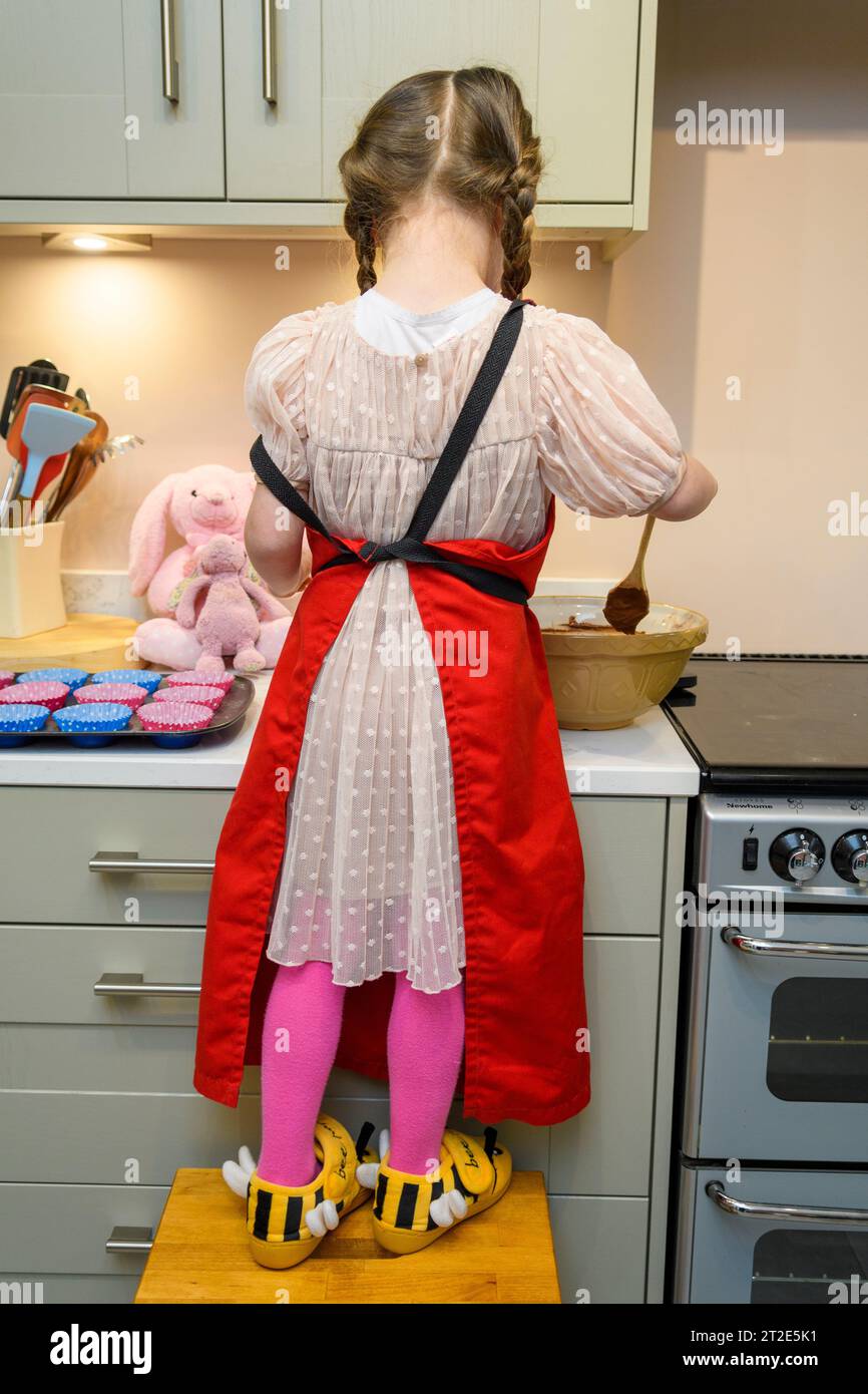 Small child standing on a stool wearing an apron in a kitchen stirring ...