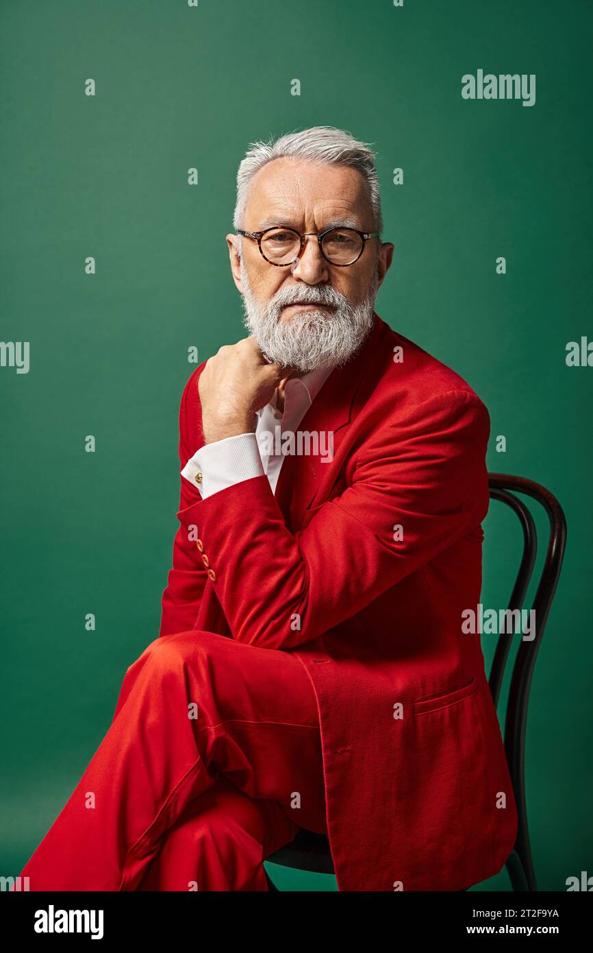 elegant handsome Santa in red suit posing on chair with his hand near ...