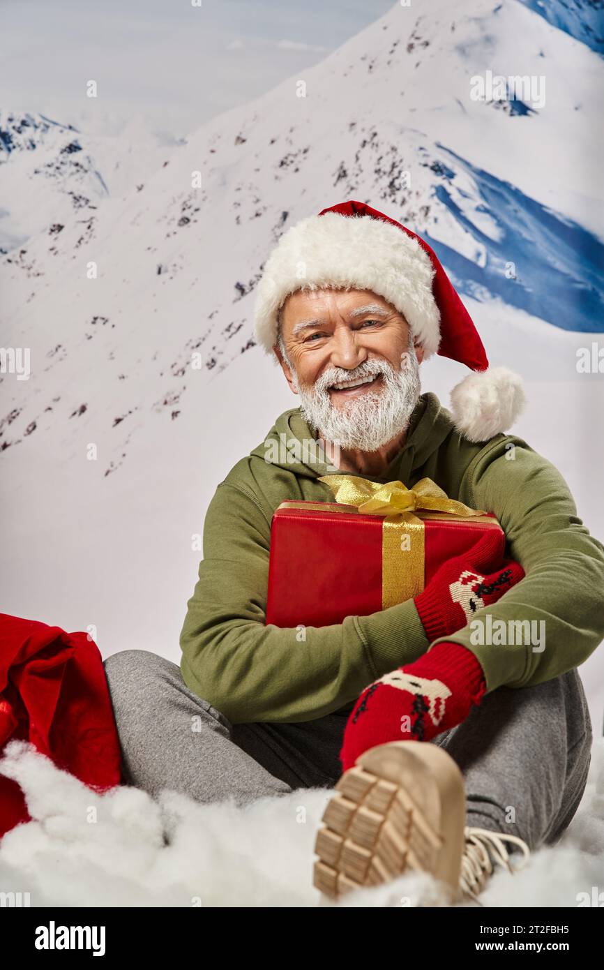 joyful white bearded man dressed as Santa sitting on snow with present ...