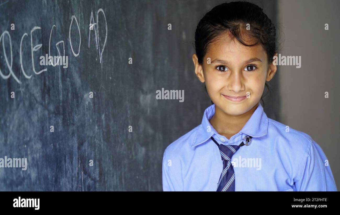 Happy Indian school girl child standing in front of black chalkboard background. Education ...