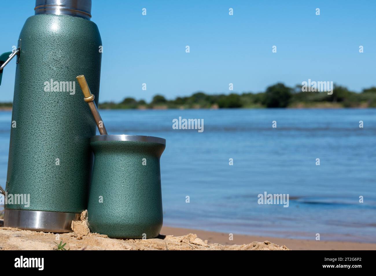 Thermos and mate, resting on sand on the bank of a river. In the ...