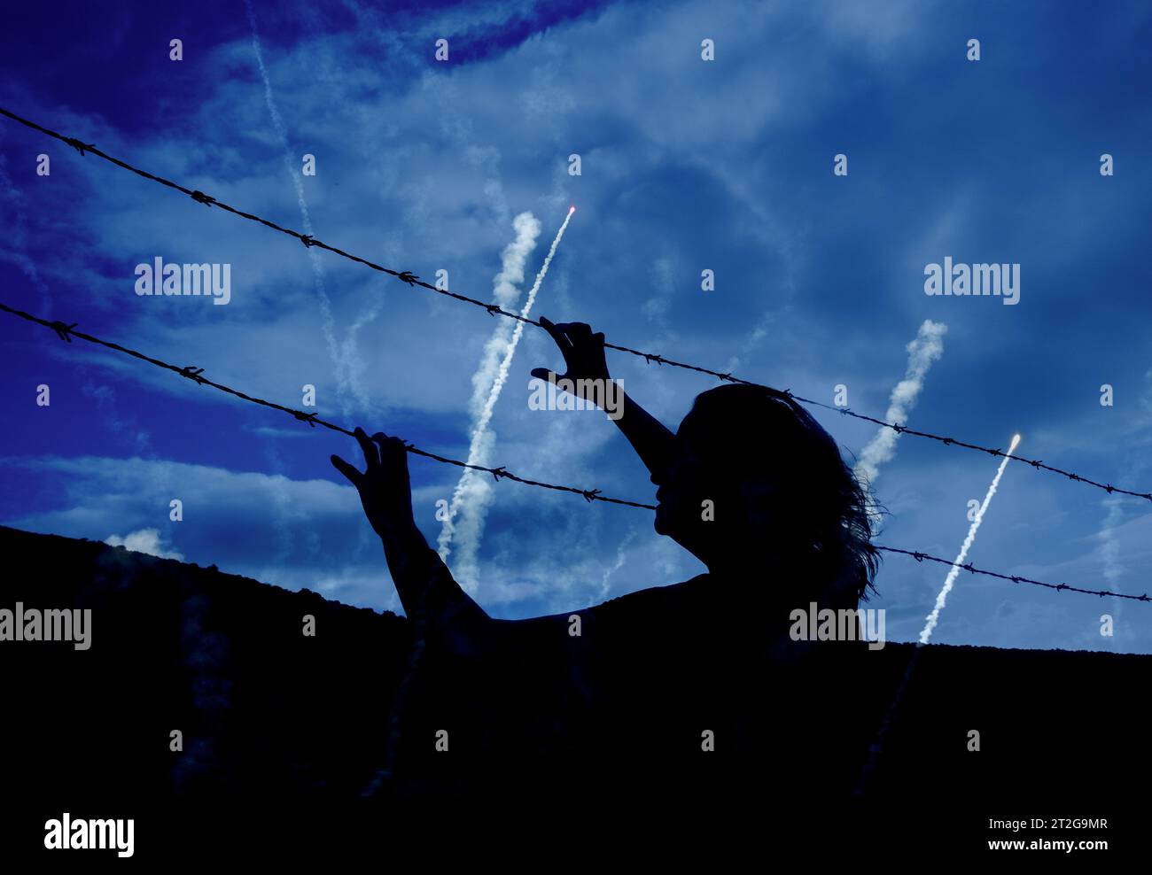 Woman looking through barbed wire fence with shelling in background ...