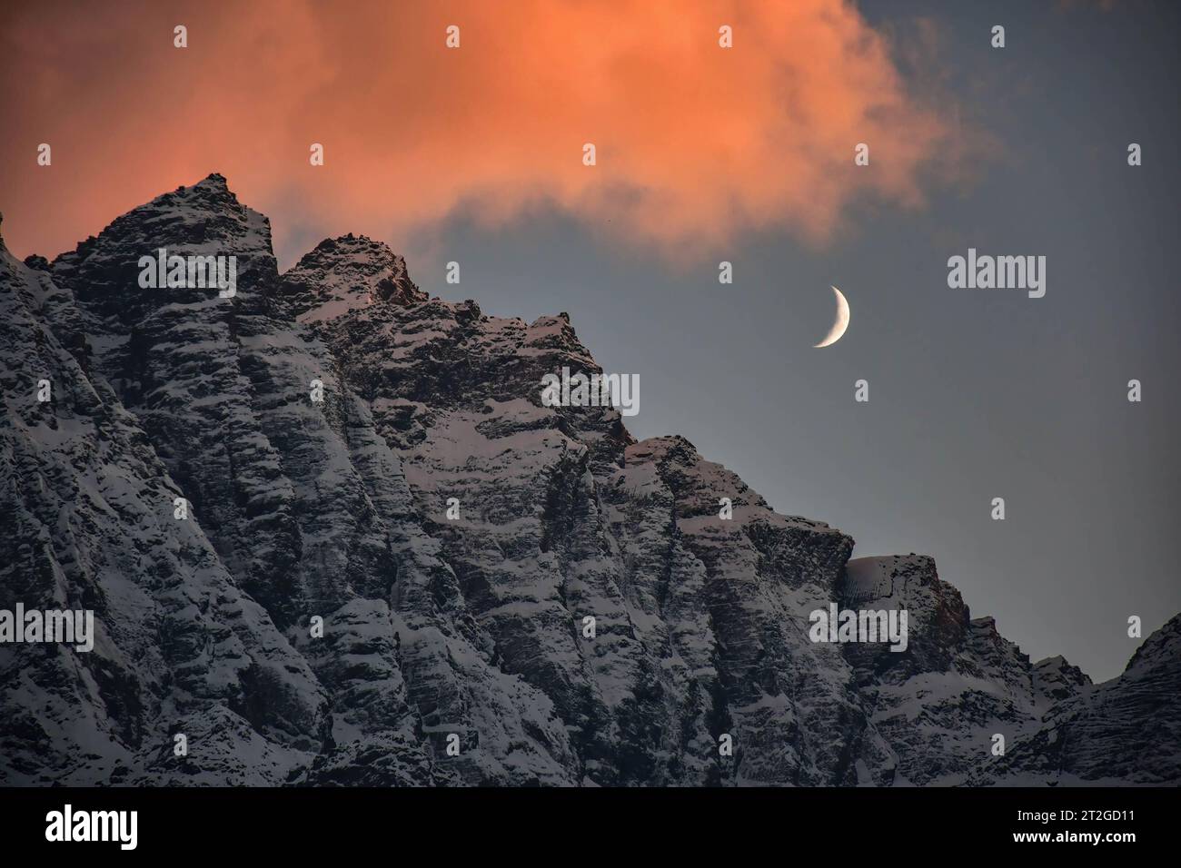 The waxing crescent moon rises over the snow covered mountains in ...