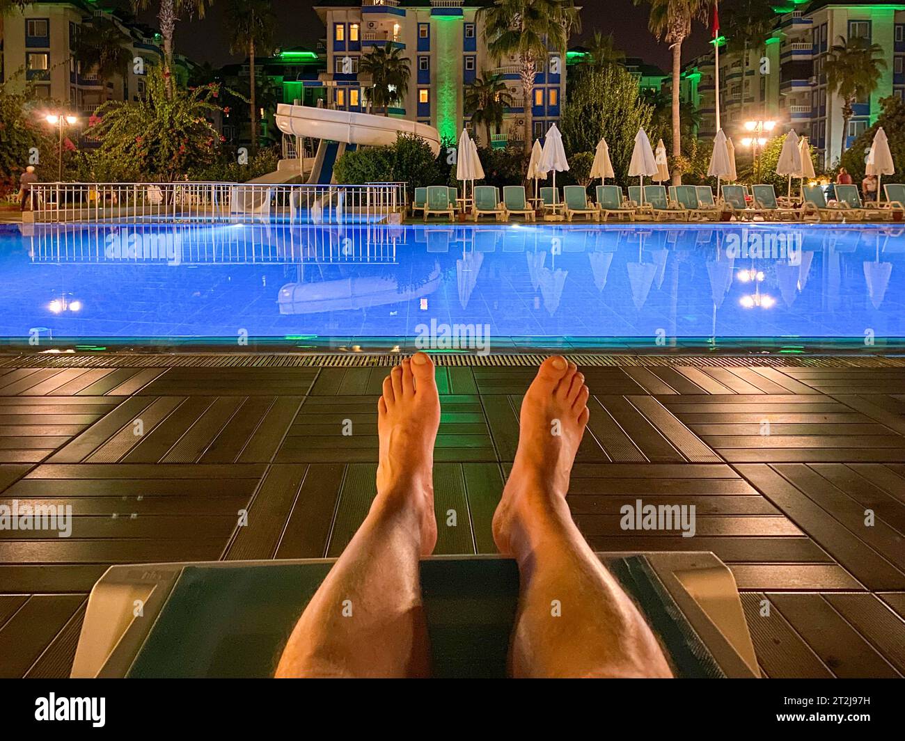 Men's feet on a sun lounger by the pool at night in a hotel in a warm ...