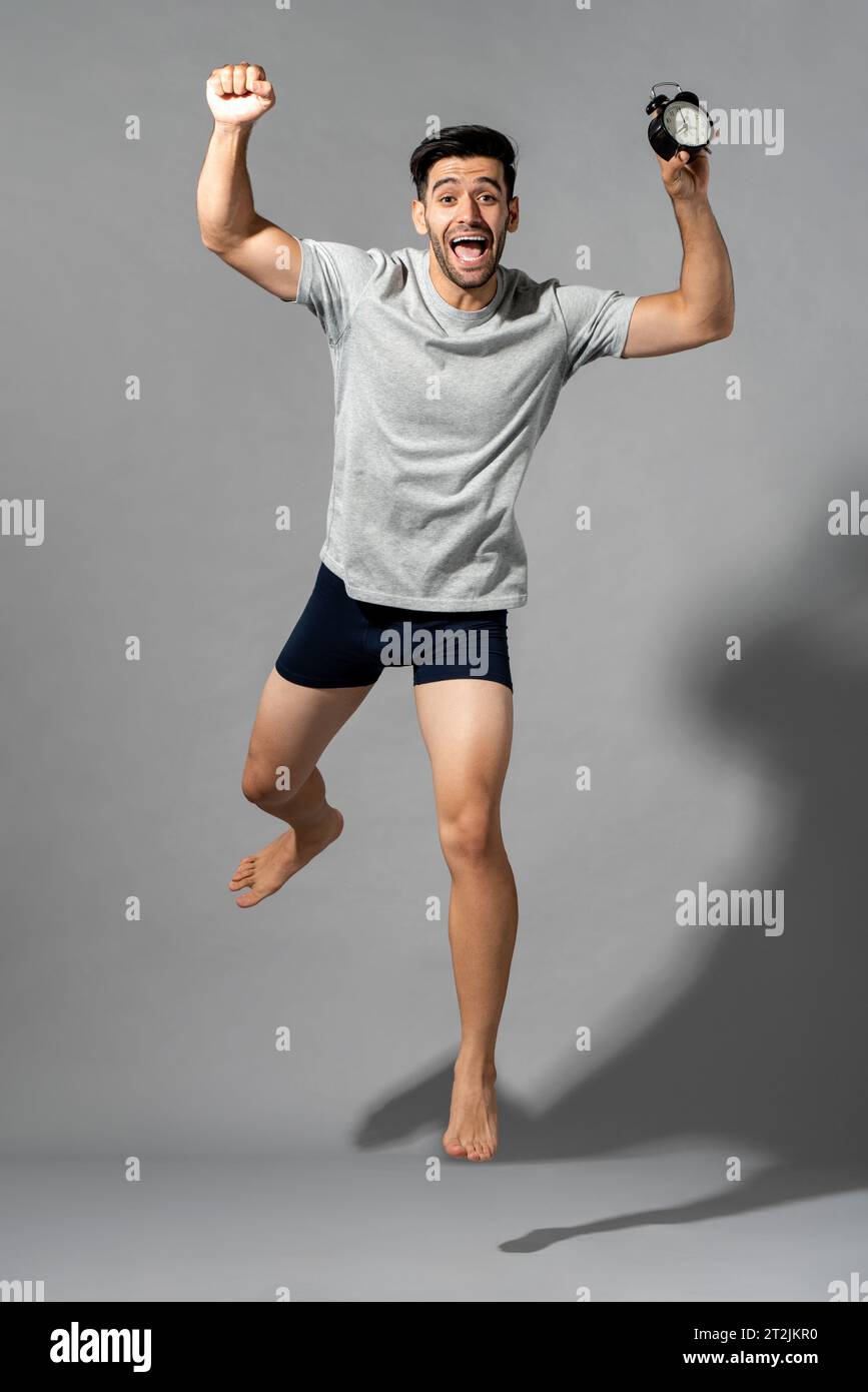 Cheerful Caucasian man with alarm clock in hand jumping on gray ...