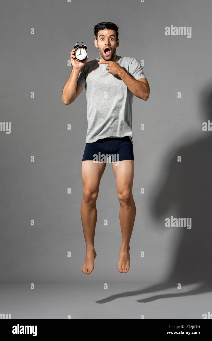 Shocked Caucasian man with alarm clock in hand jumping after waking up ...