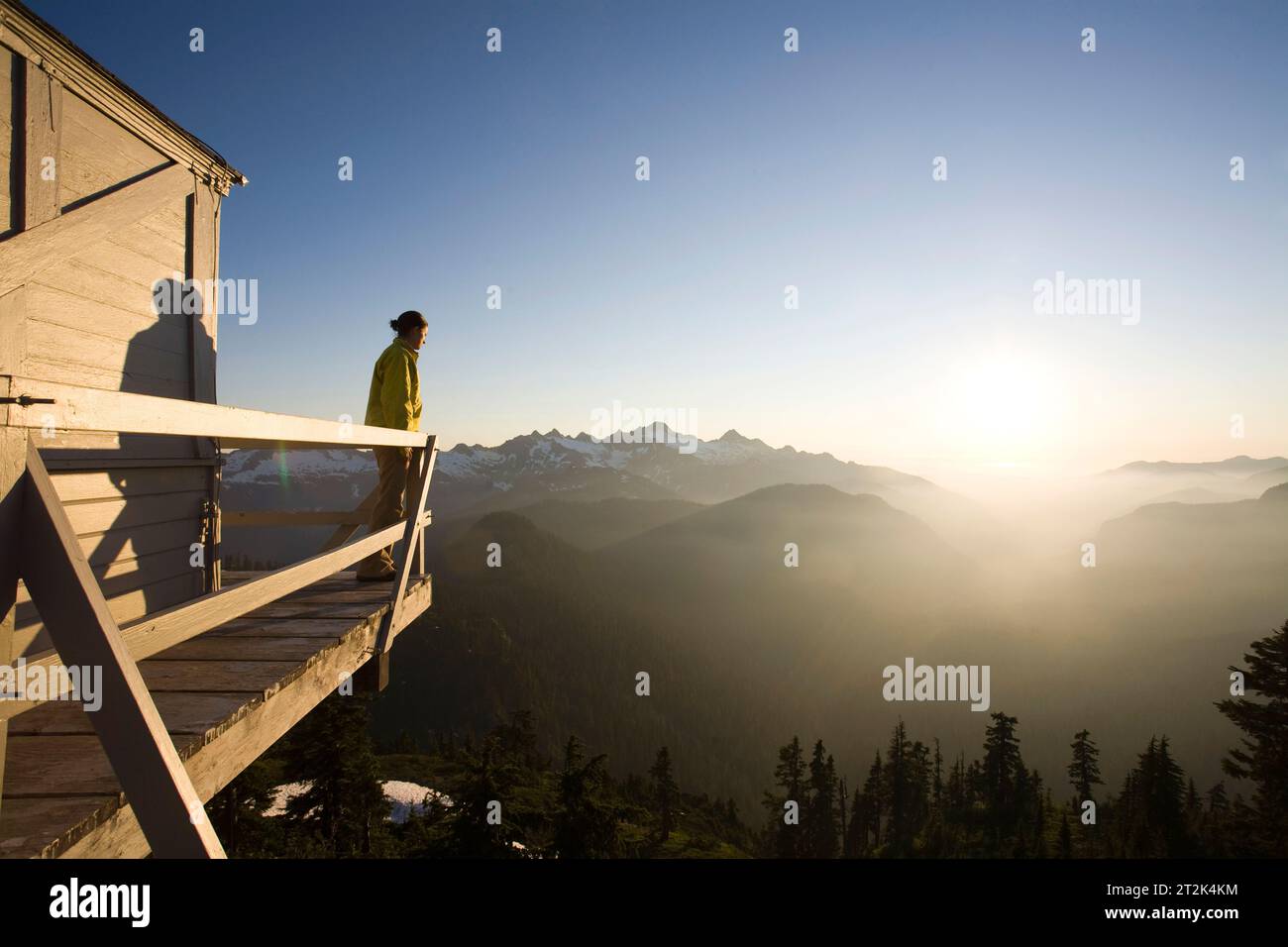 A young woman stands on the porch of a fire lookout tower overlooking ...