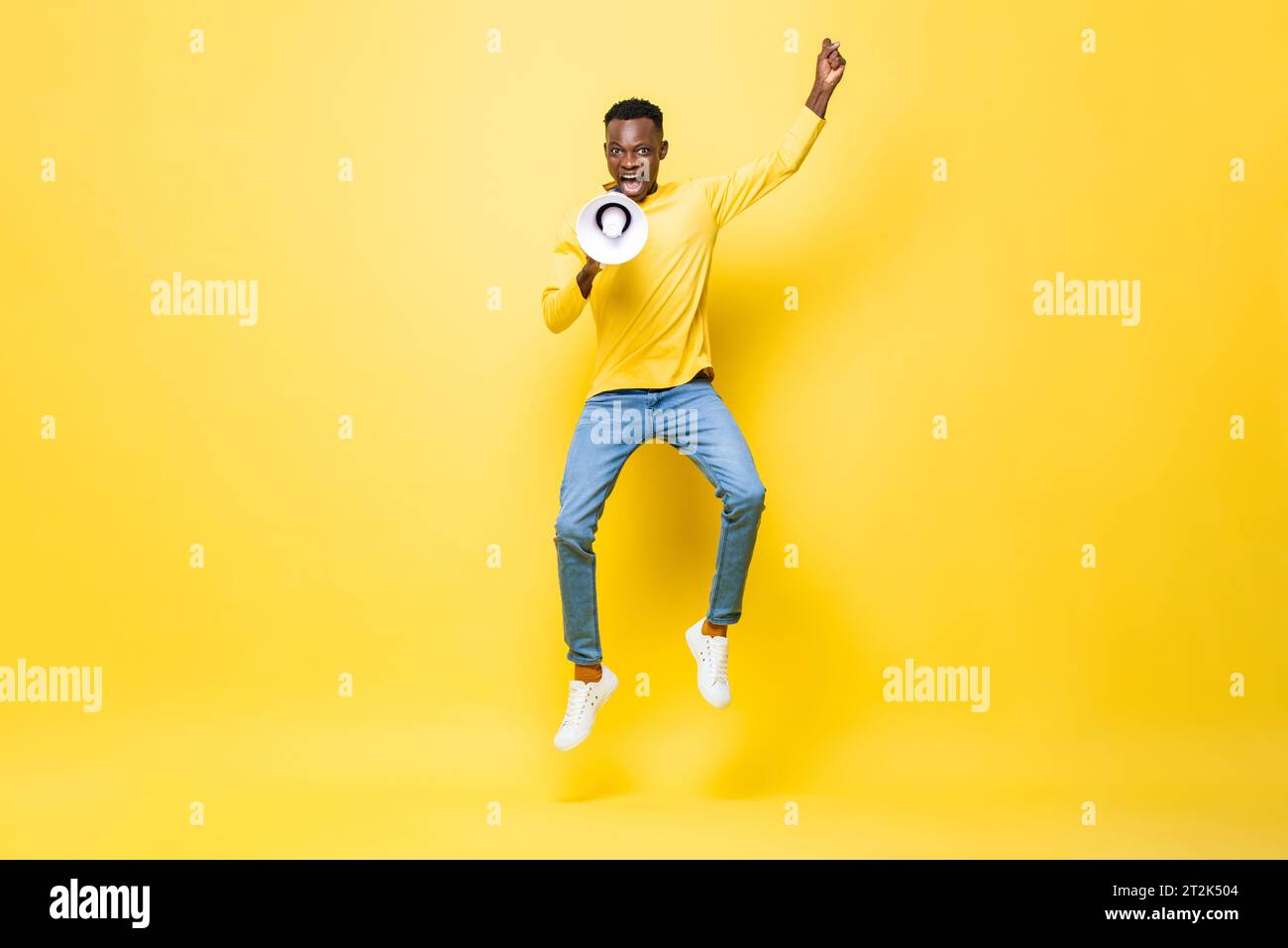 Powerful energetic young African man jumping and yelling on megaphone in studio yellow color ...