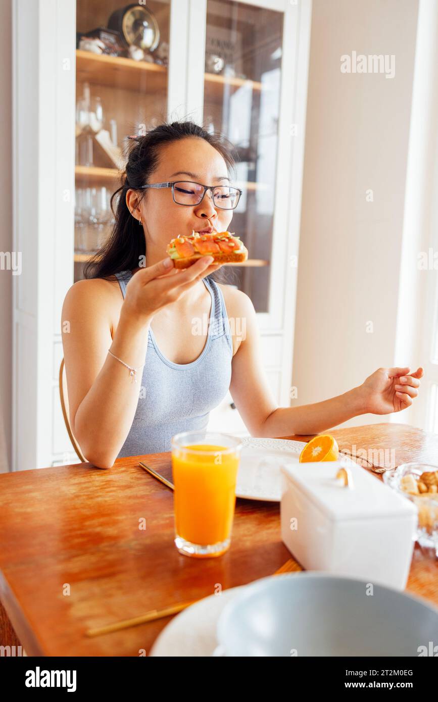 Lovely Asian woman in glasses and casual clothes eating sandwich with ...
