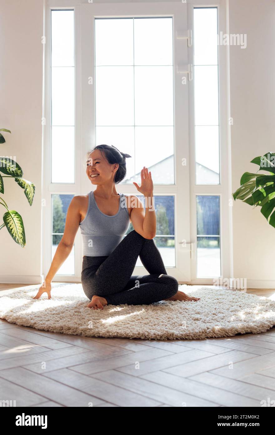 Cute young Asian woman in fitness suit smiles and does yoga in living ...