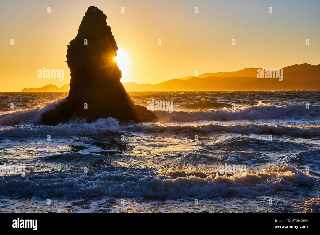 Tall massive boulder jutting out of ocean waves with golden sun poking ...