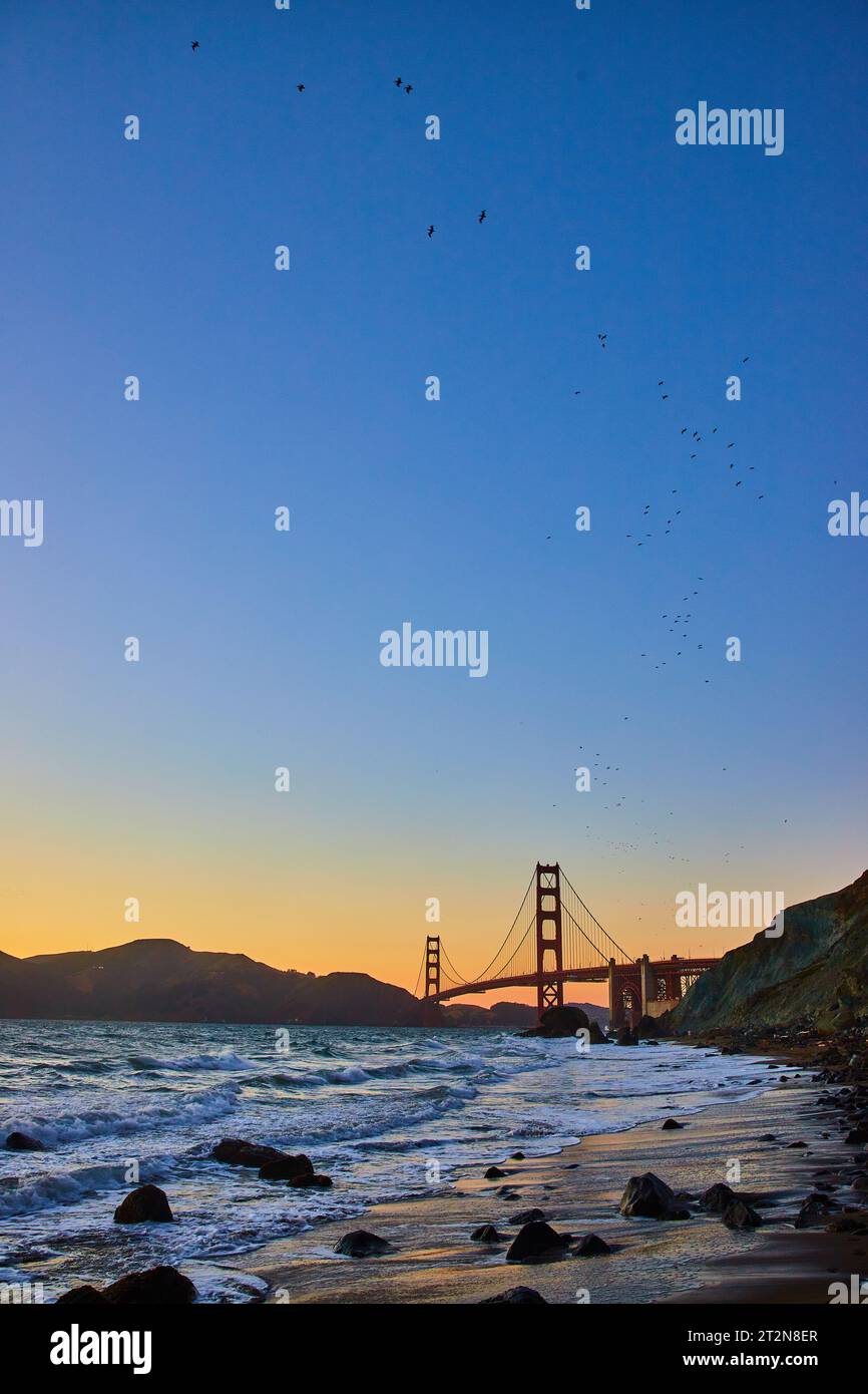 Sunset sky above glowing blue waves and Golden Gate Bridge along sandy ...