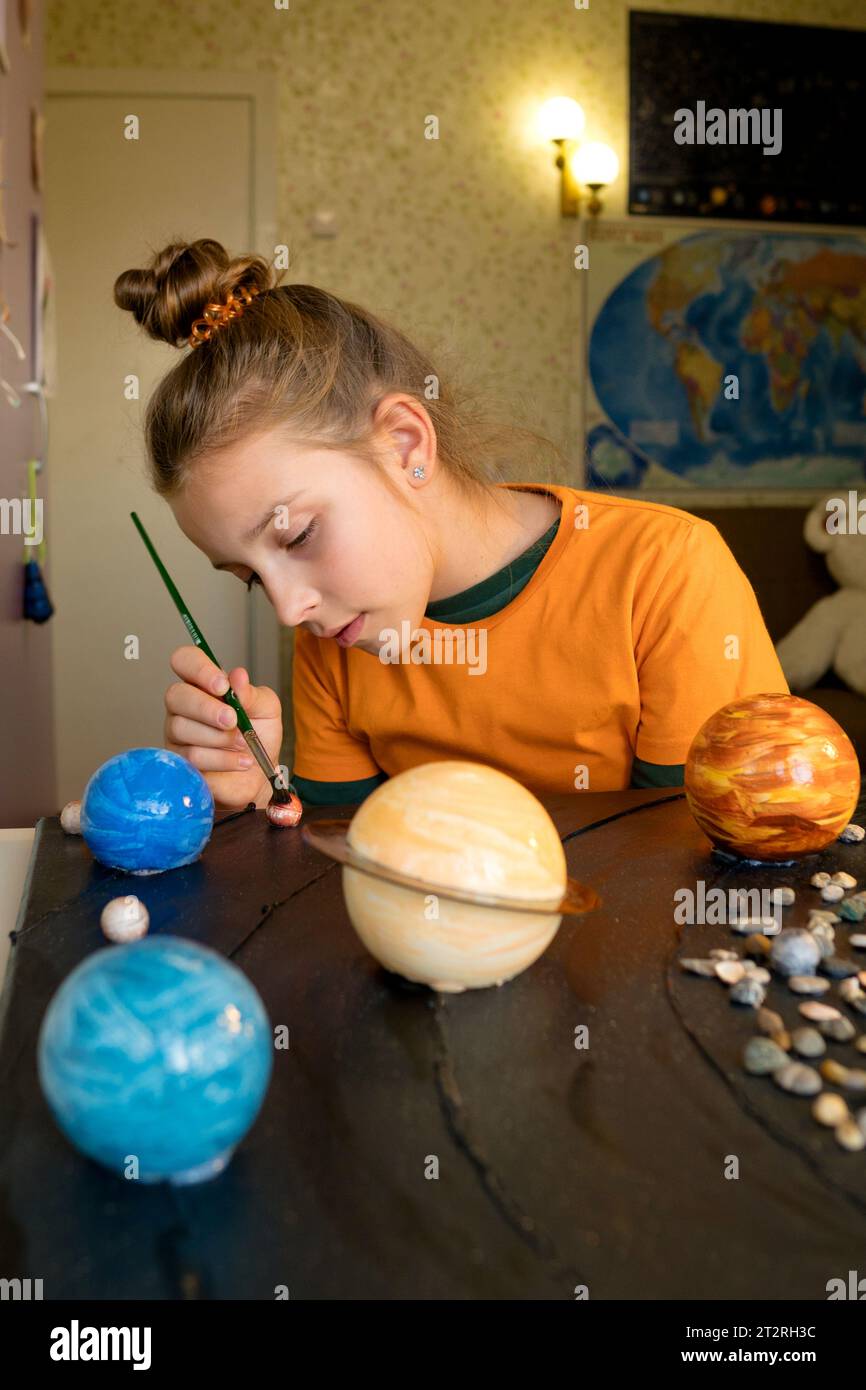 Focused young girl delicately painting planetary models with precision, set against a backdrop ...