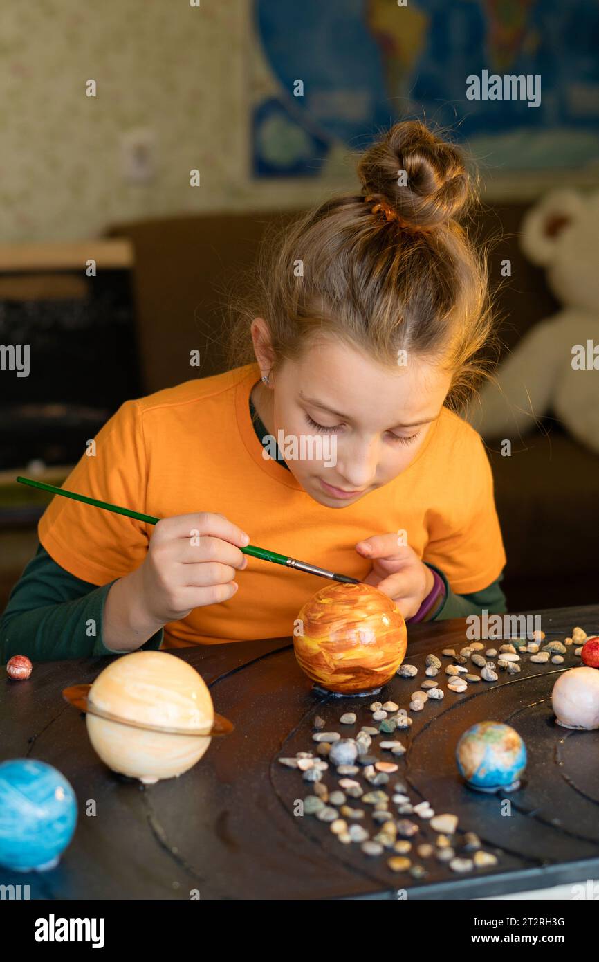 Focused young girl delicately painting planetary models with precision, set against a backdrop ...