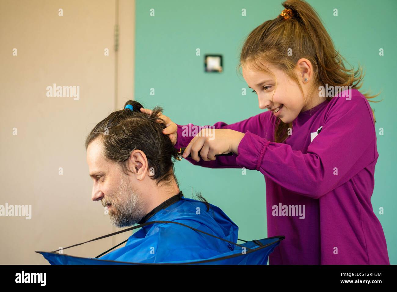 Joyful girl playfully cuts her father's hair at home, creating a bond ...