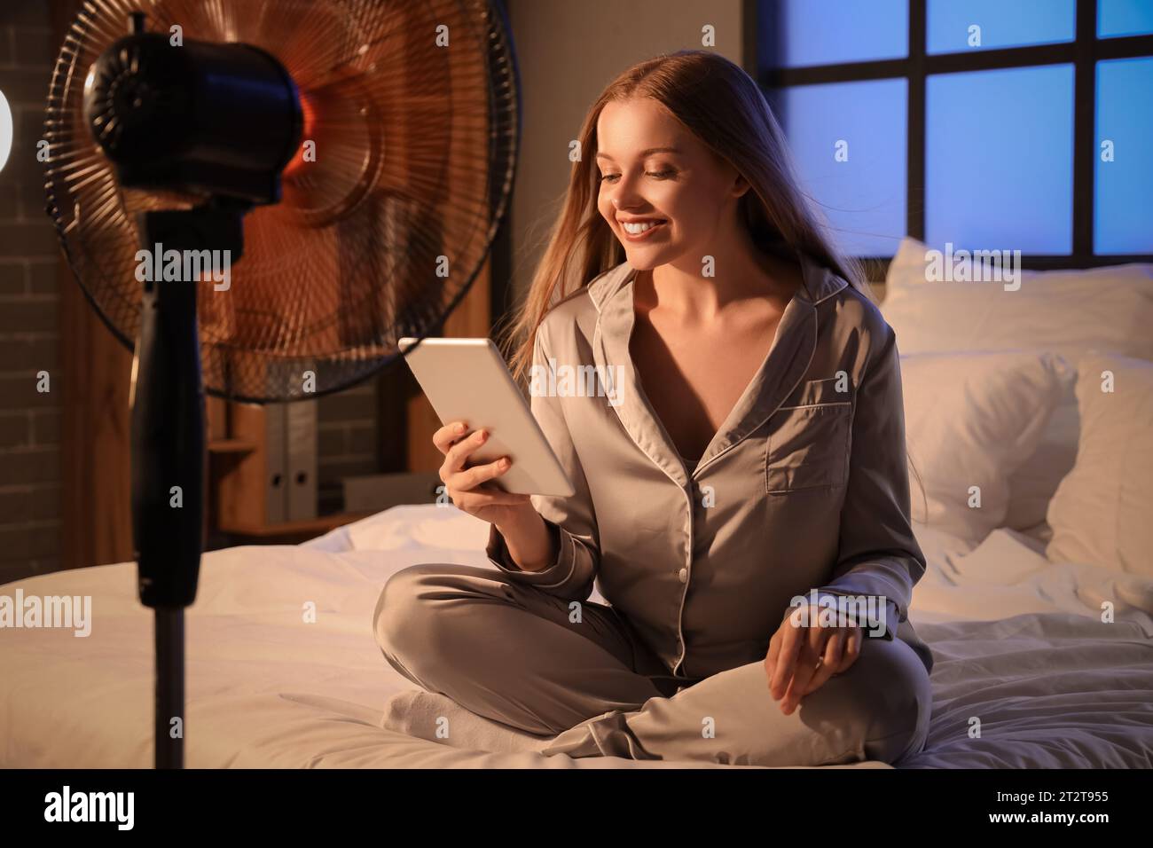 Young woman with blowing electric fan using tablet computer in bedroom ...
