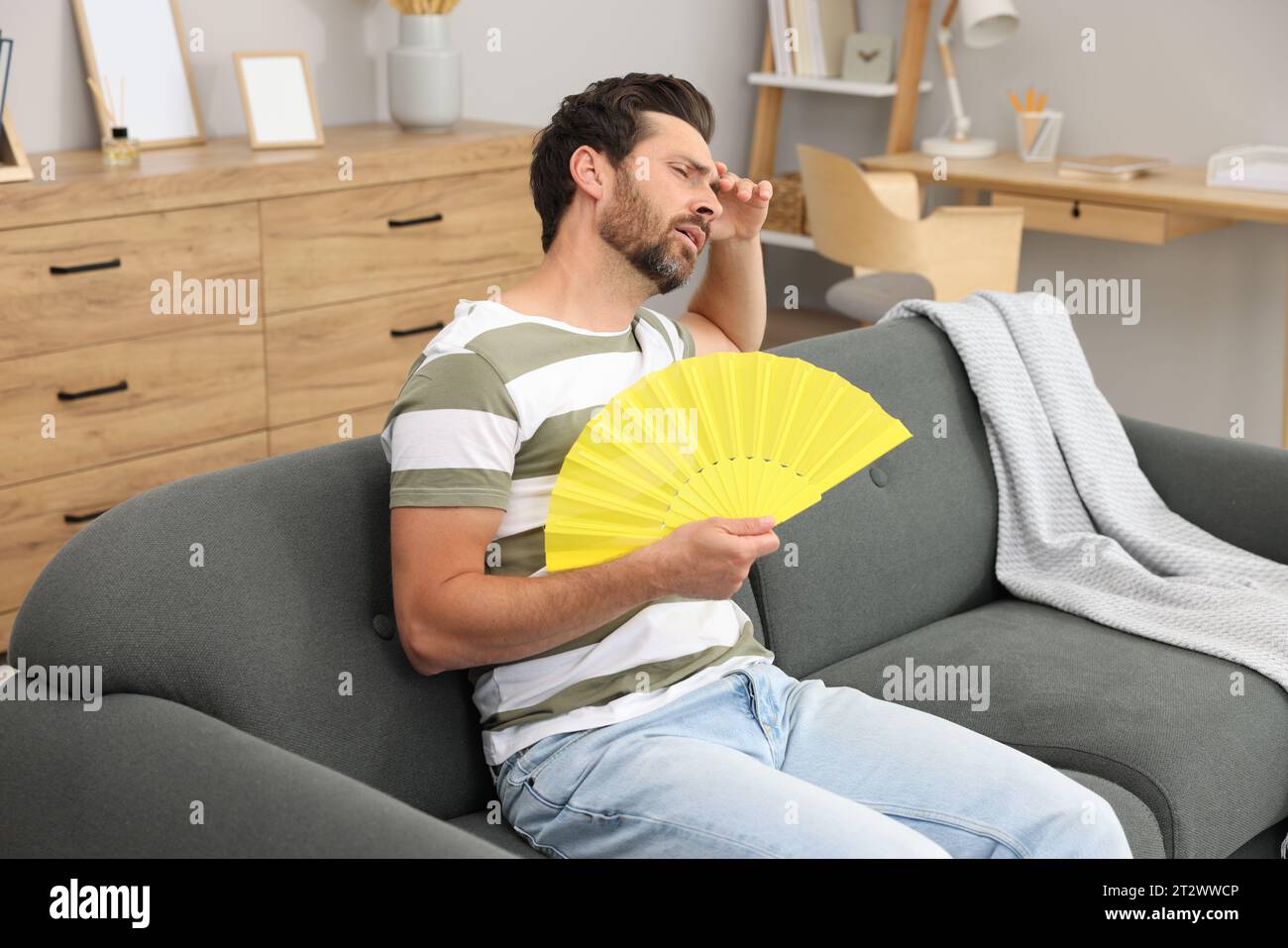 Bearded man waving yellow hand fan to cool himself on sofa at home ...