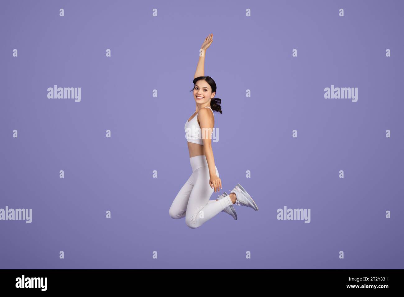 Joyful woman jumping, wearing white sports outfit, purple background ...