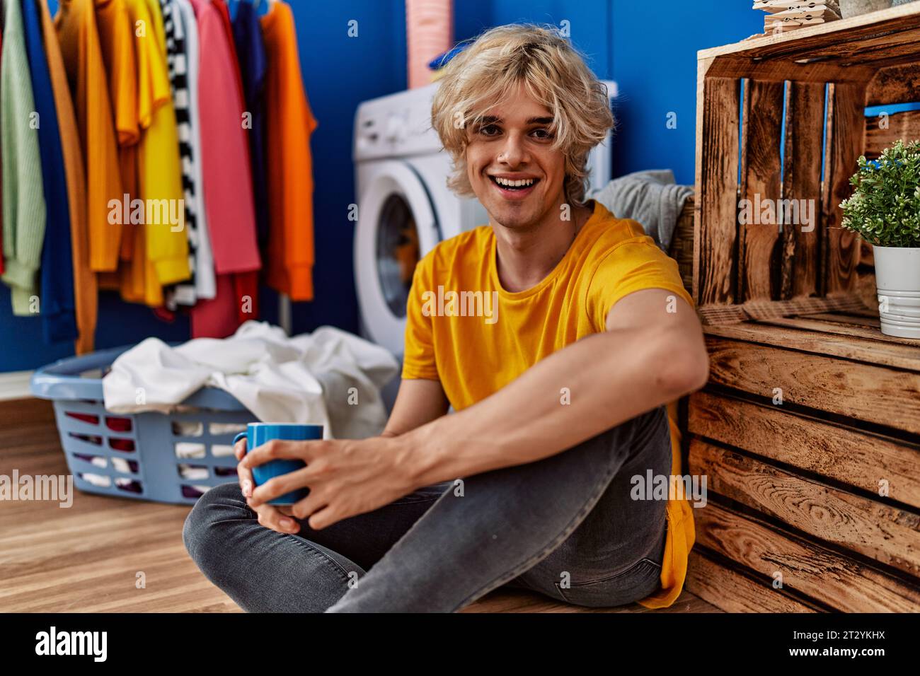 Young blond man drinking coffee waiting for washing machine at laundry ...