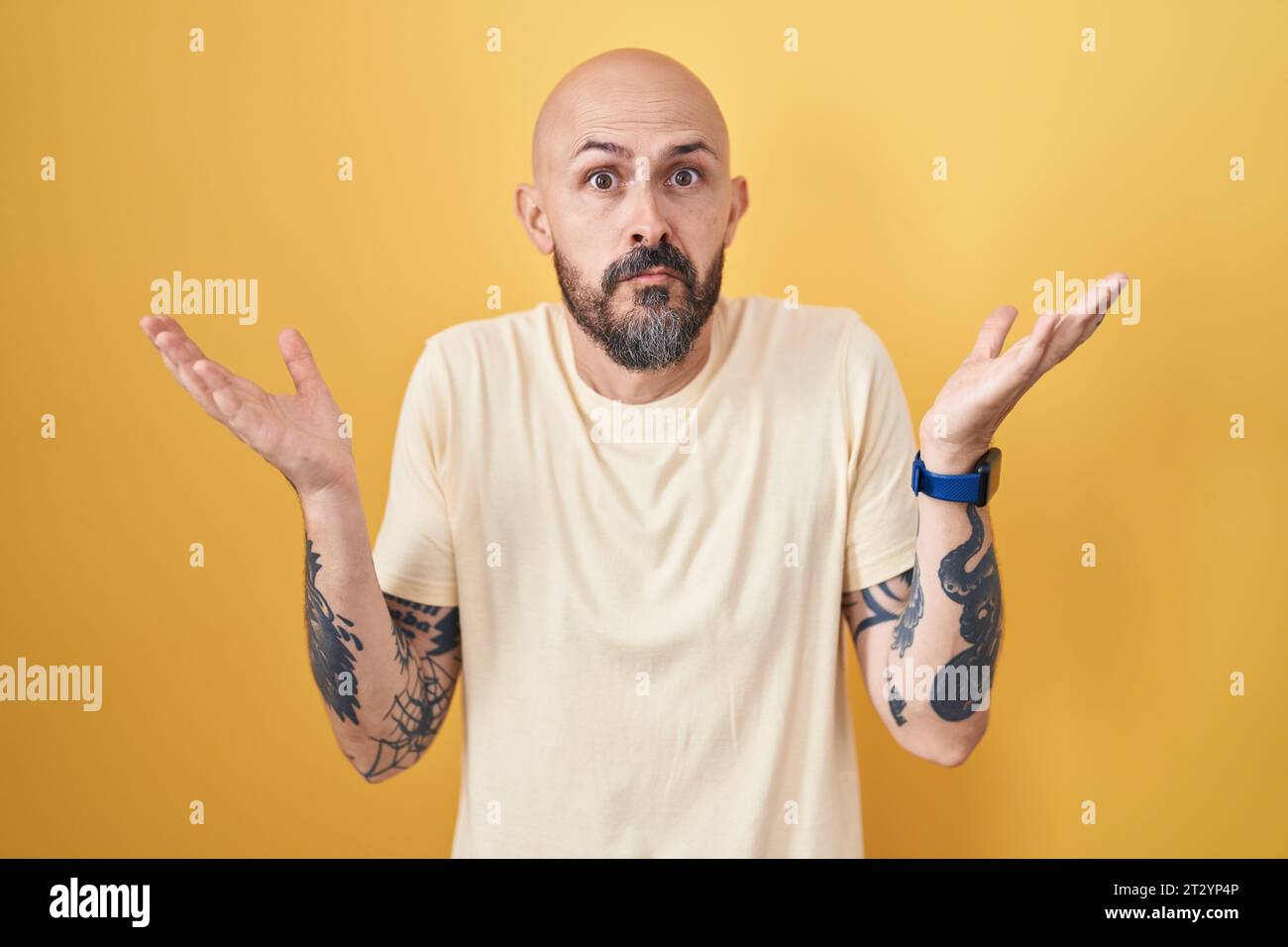 Hispanic man with tattoos standing over yellow background clueless and ...