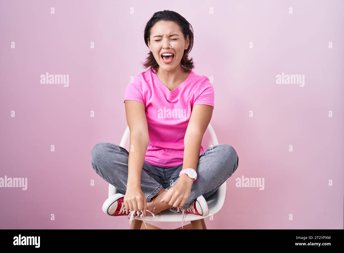 Hispanic young woman sitting on chair over pink background angry and ...