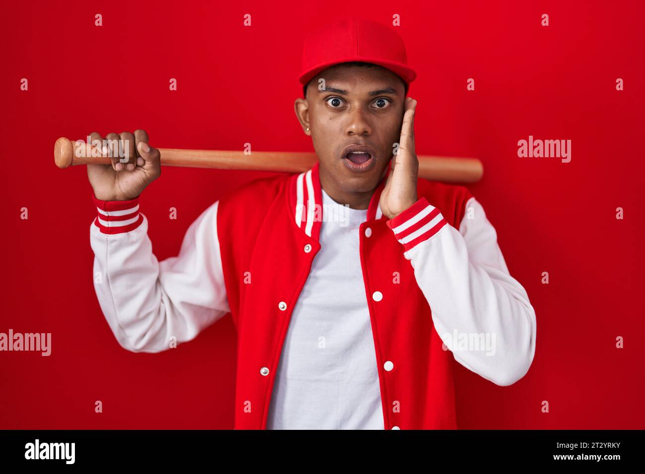Young hispanic man playing baseball holding bat afraid and shocked ...