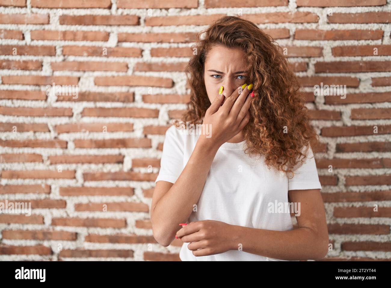 Young caucasian woman standing over bricks wall background smelling ...