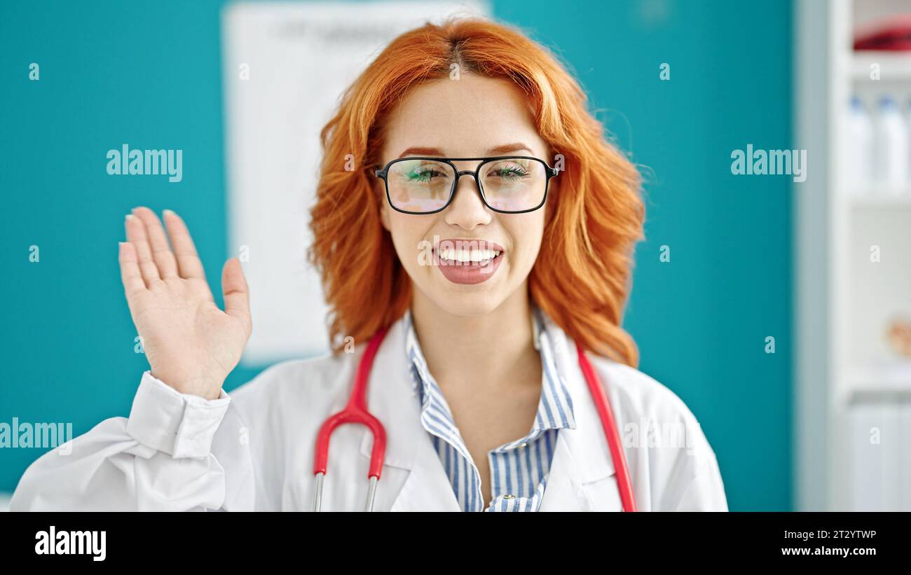 Young redhead woman doctor smiling confident saying hello with hand at ...