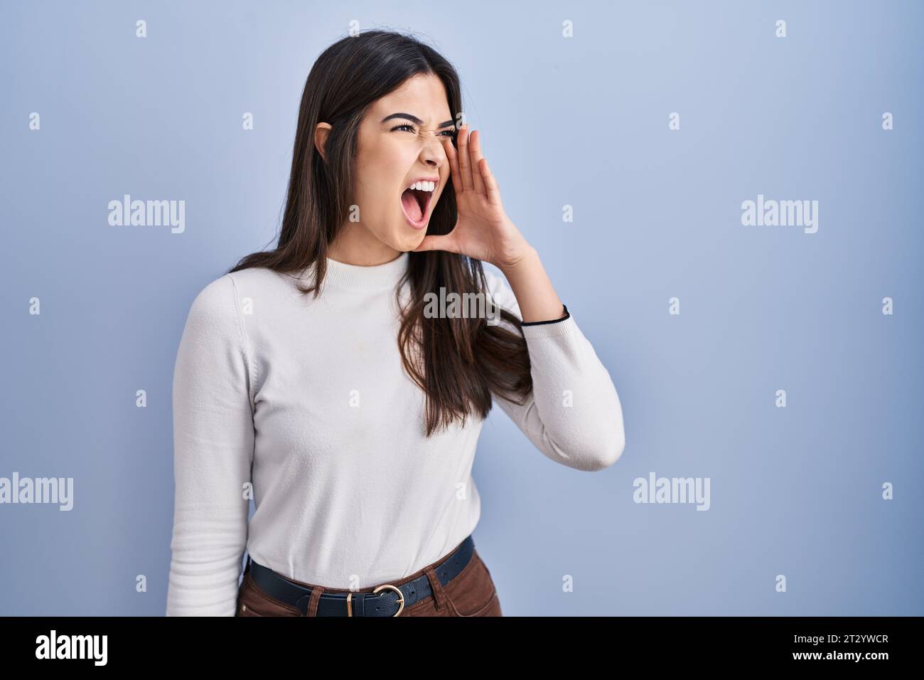 Young brunette woman standing over blue background shouting and ...