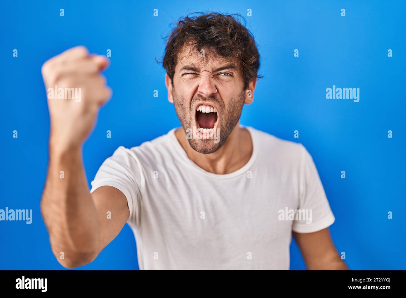Hispanic young man standing over blue background angry and mad raising ...