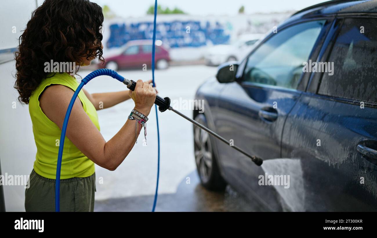 Middle age hispanic woman washing car with pressure washer at car wash ...