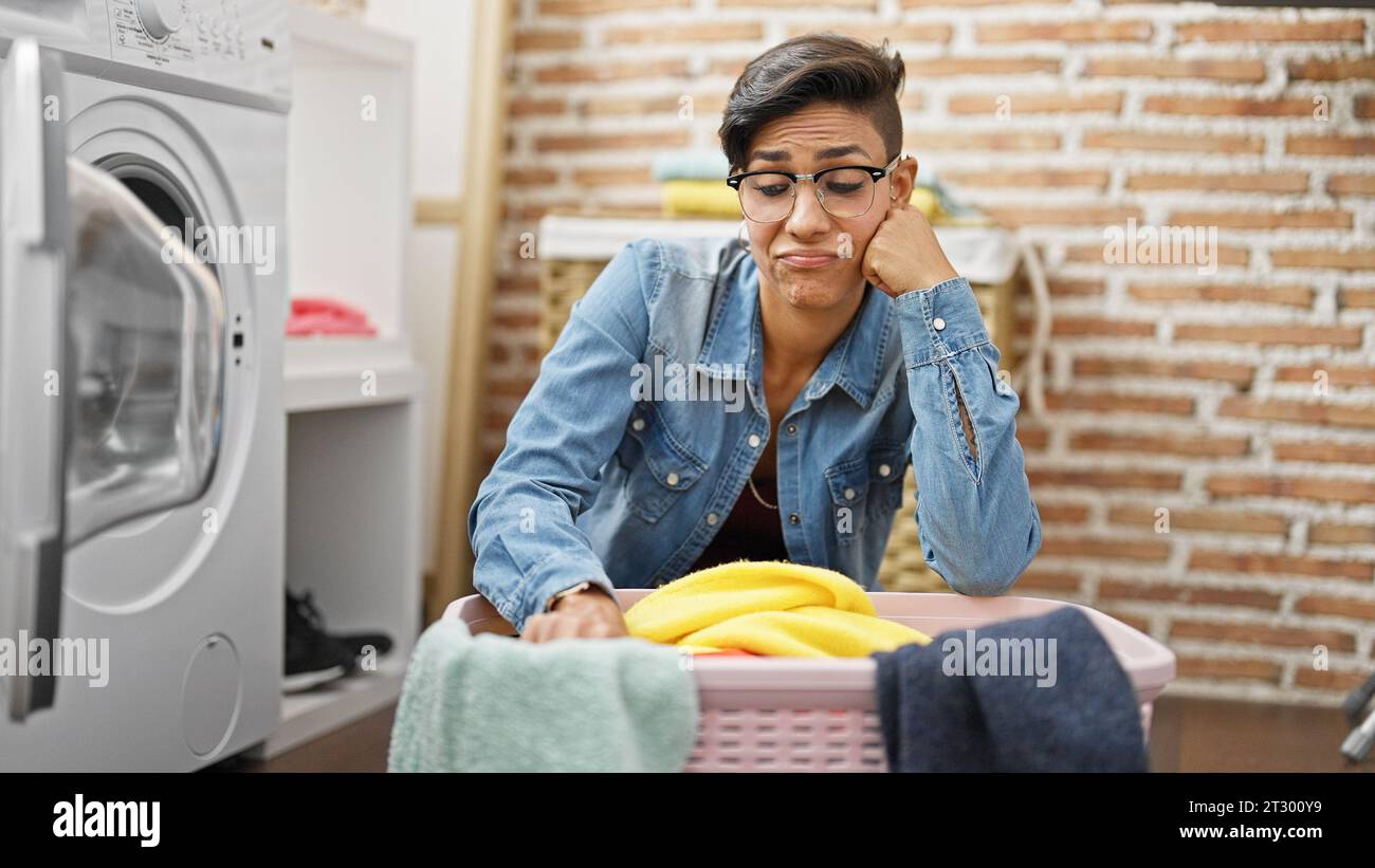 Young beautiful hispanic woman leaning on basket with clothes sadness ...