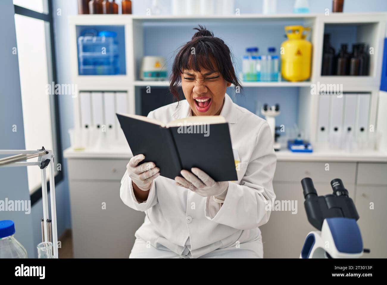 Hispanic woman working at scientist laboratory reading a book angry and mad screaming frustrated ...