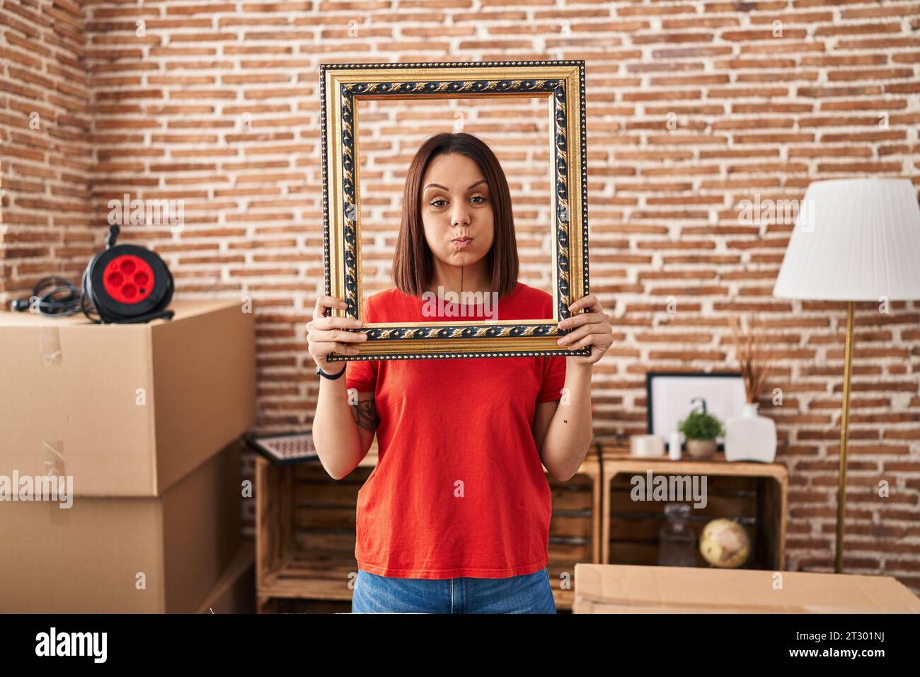 Young hispanic woman at new home holding empty frame puffing cheeks ...