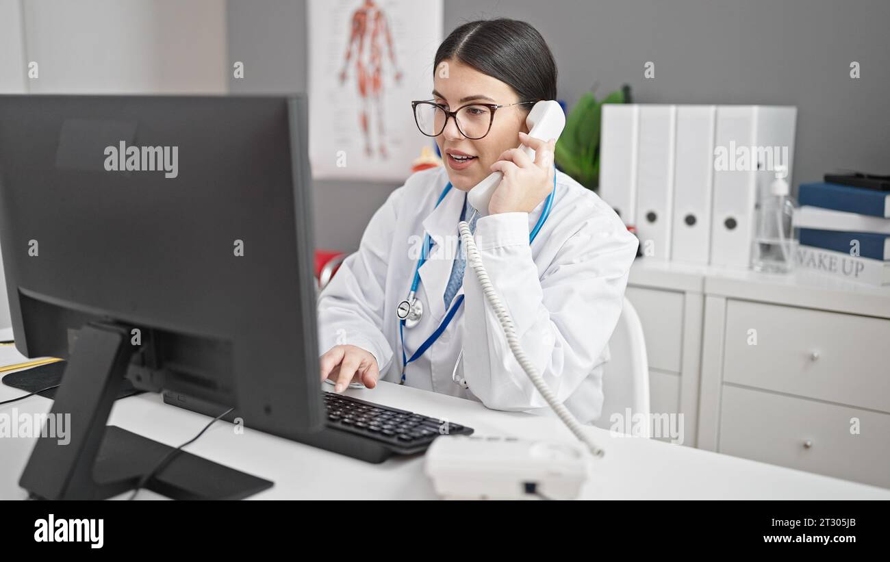 Young hispanic woman doctor using computer speaking on telephone at ...