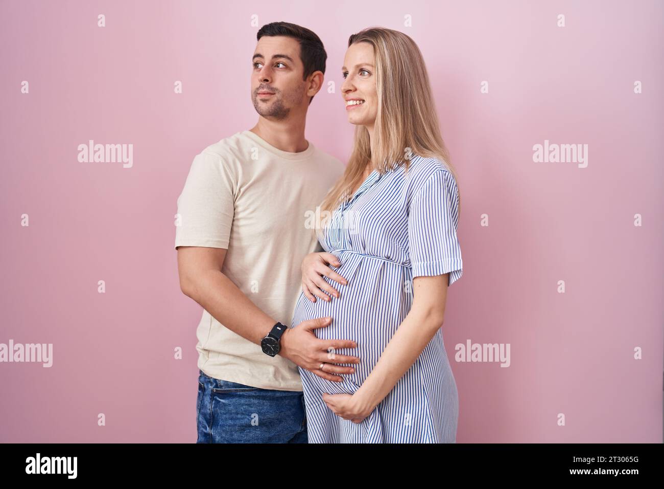 Young couple expecting a baby standing over pink background looking to ...