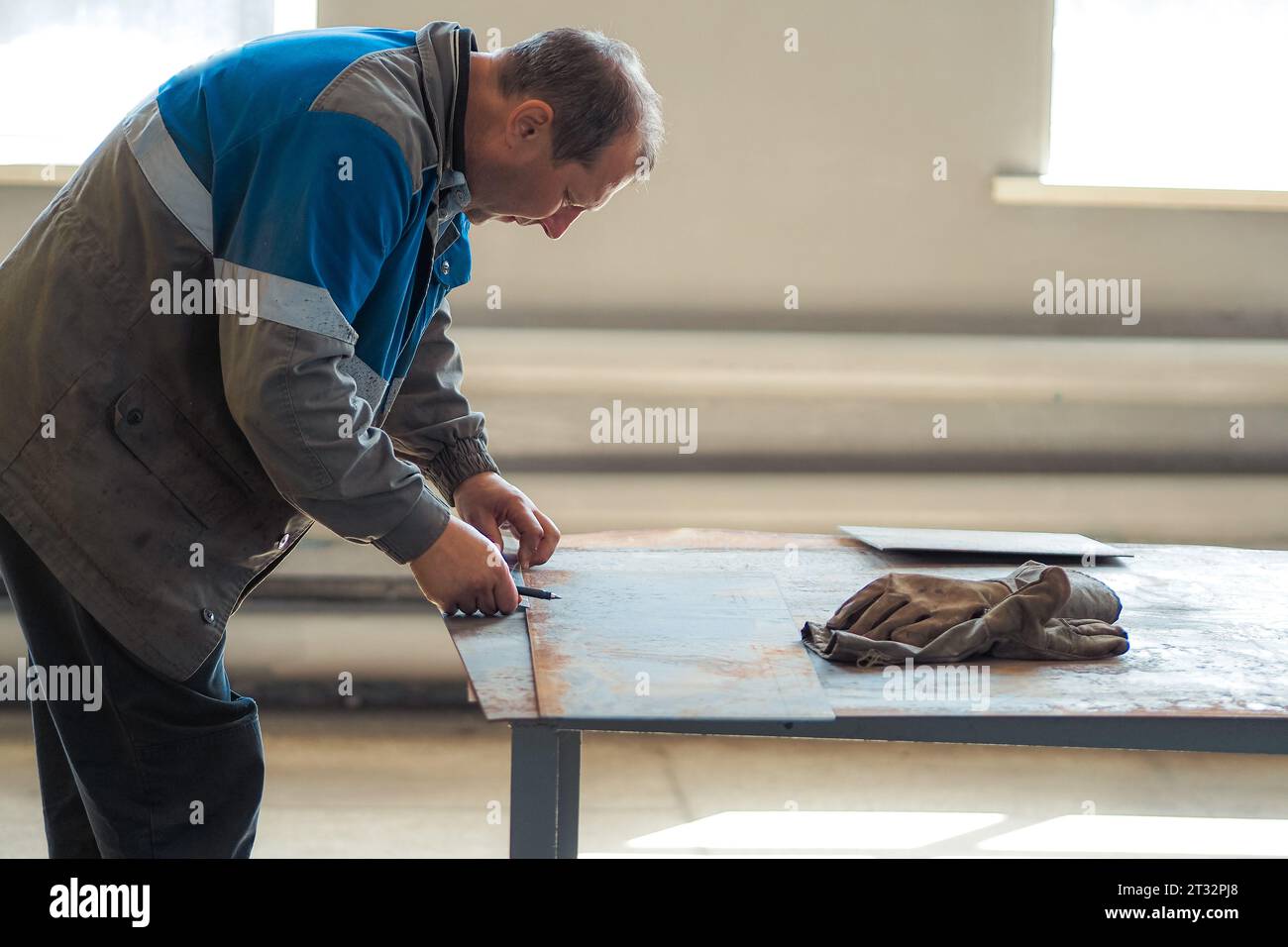 Worker in overalls works in workshop at workbench. Man marks metal ...