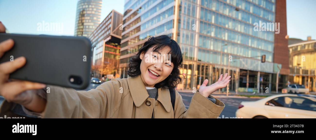 Smiling korean girl showing city, taking selfie in front of buildings ...