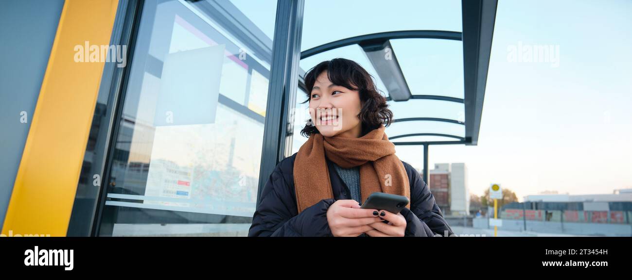 Cute smiling asian girl standing on bus stop, holding smartphone ...