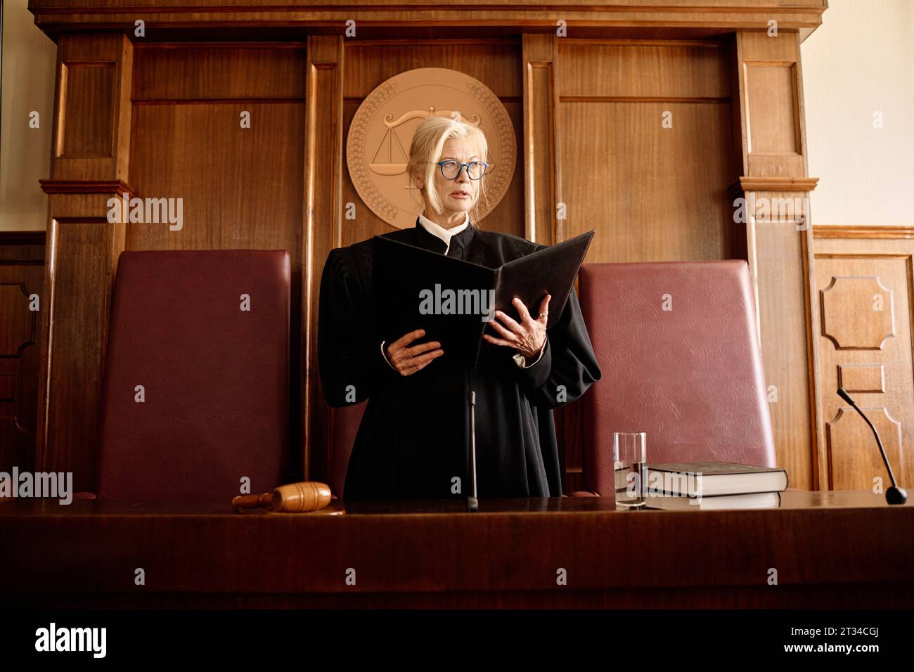 Confident mature blond female judge with open folder standing by desk ...