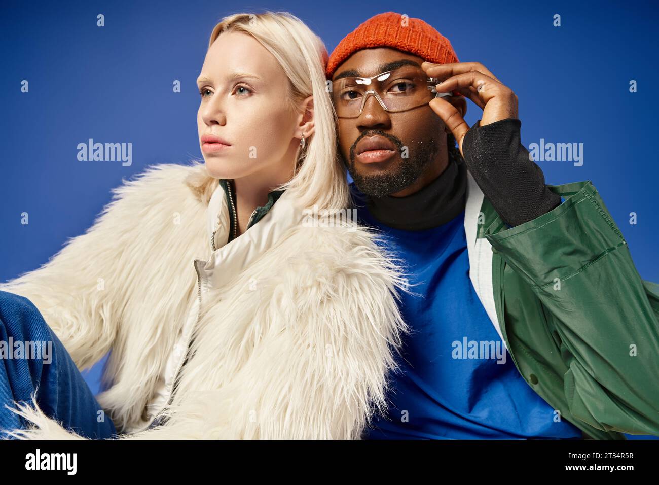 multicultural couple in winter attire sitting together on blue backdrop ...