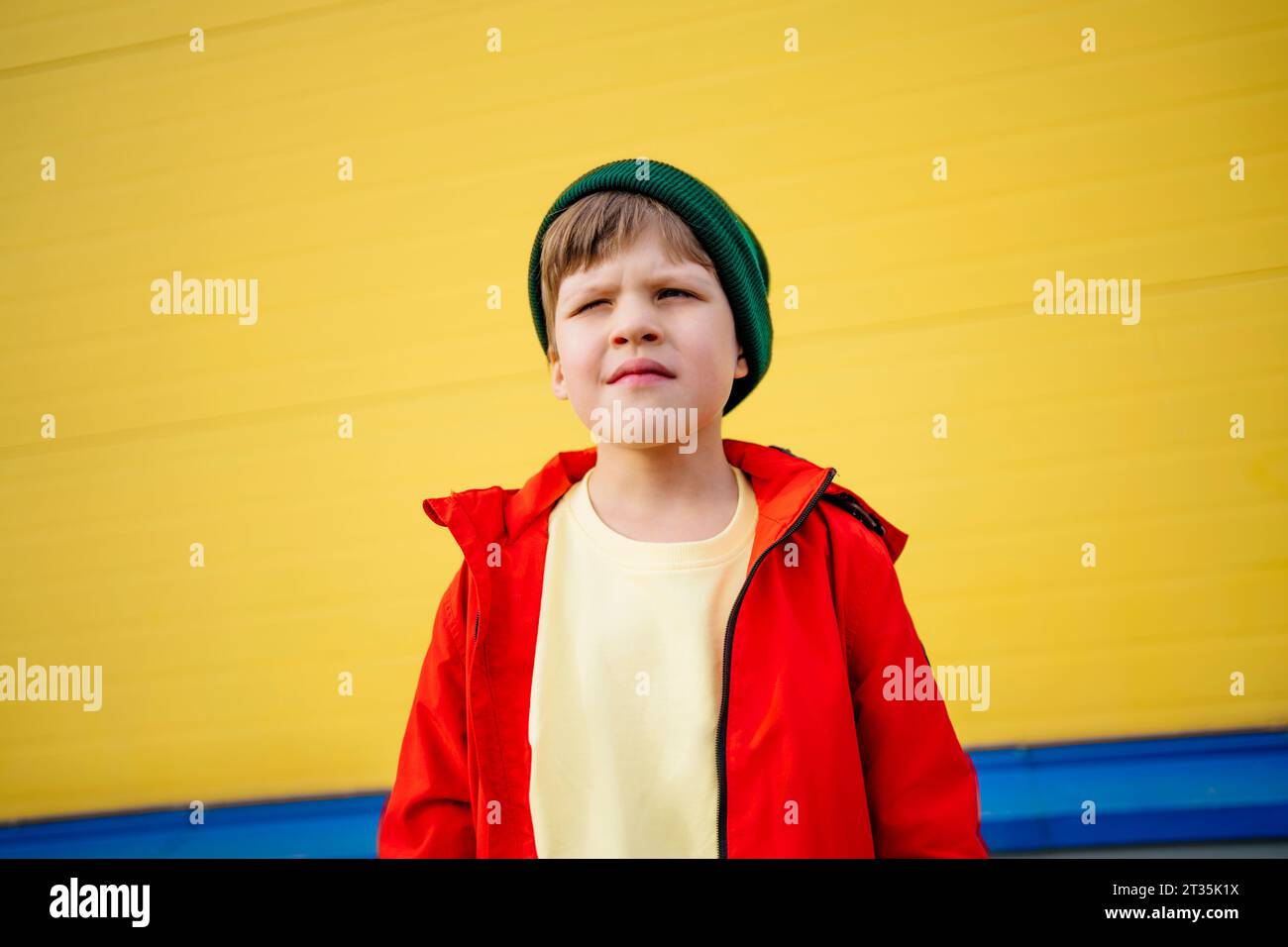Boy wearing red jacket and knit hat standing in front of yellow wall ...