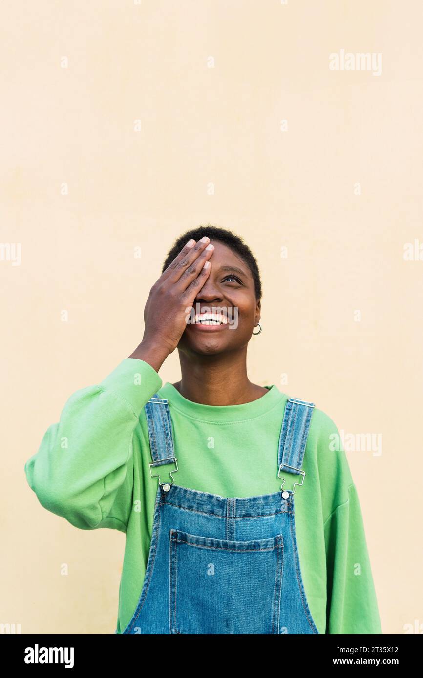 Happy young woman covering eye with hand standing in front of beige ...