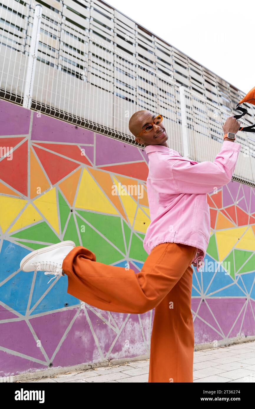 Smiling non-binary person standing on one leg in front of colorful wall ...