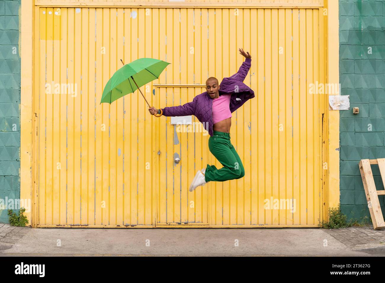 Non-binary person jumping with umbrella in front of yellow shutter door ...