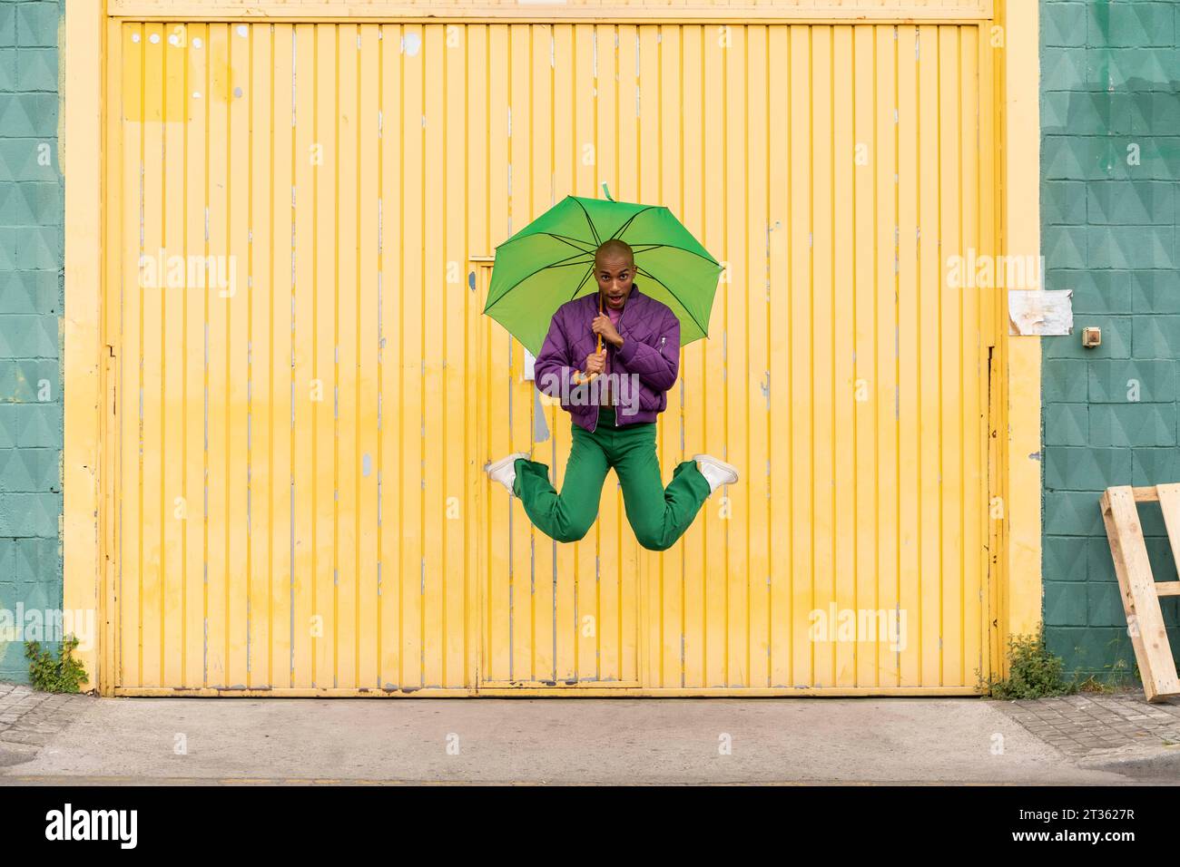 Non-binary person jumping with green umbrella in front of yellow ...