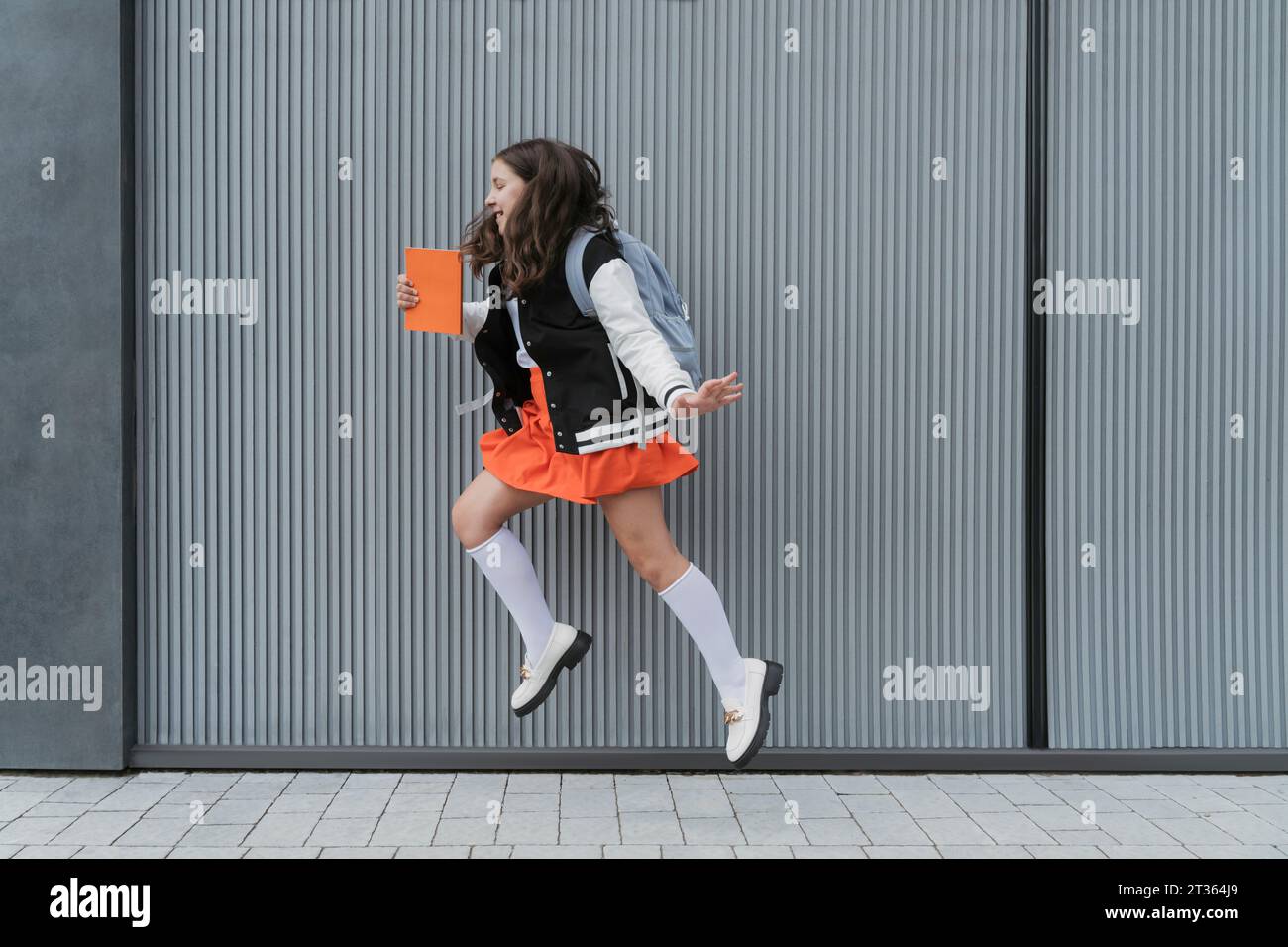 Smiling schoolgirl holding book and jumping in front of wall Stock ...