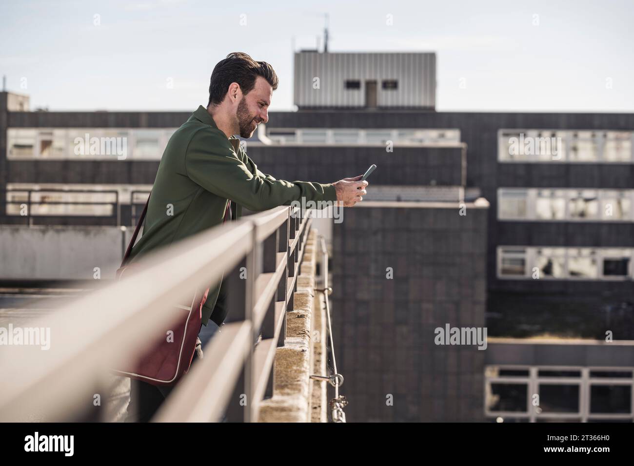 Happy man using smart phone and leaning on railing in front of building ...